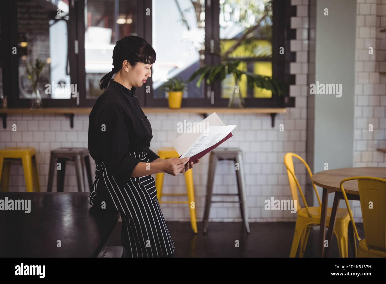 Side view of young waitress reading menu while leaning on table at ...