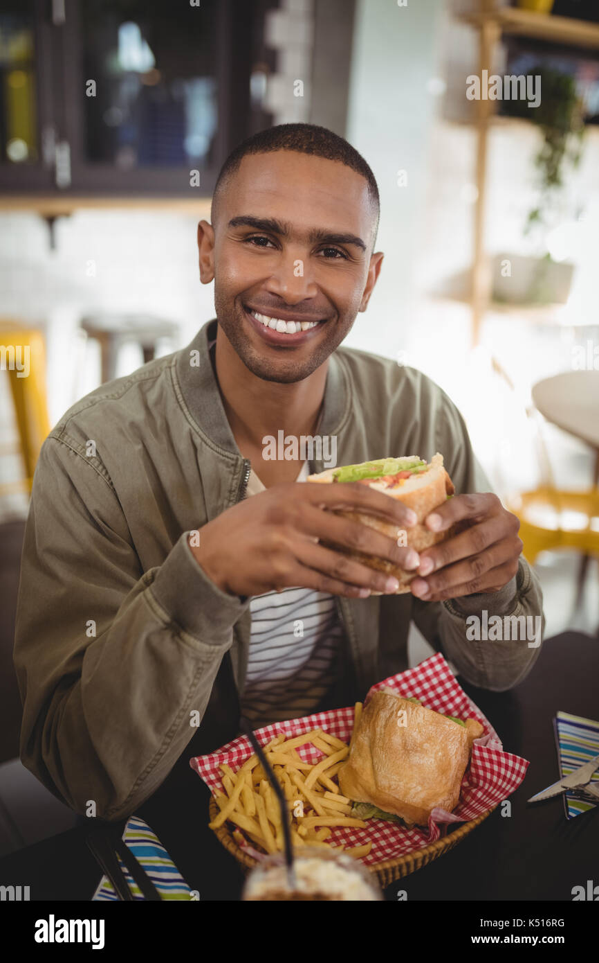 Portrait of smiling young man eating fresh sandwich while sitting at ...