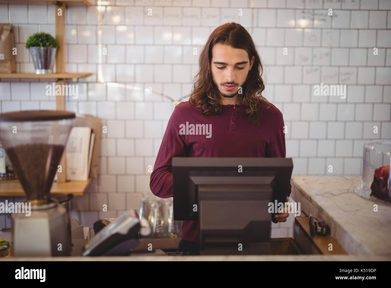 Young waiter with long hair using cash register against wall at coffee ...