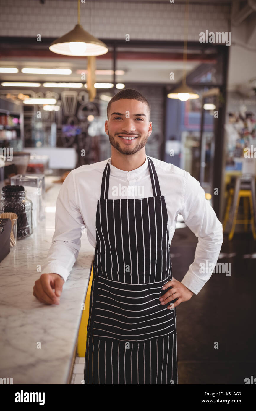 Portrait of smiling waiter standing with hand on hip by counter in ...