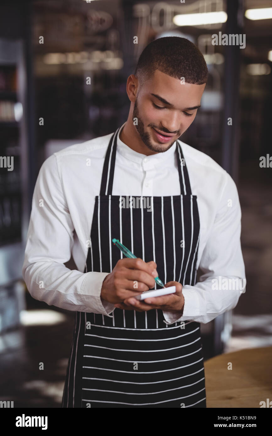 Young waiter writing in notepad while standing at coffee shop Stock ...