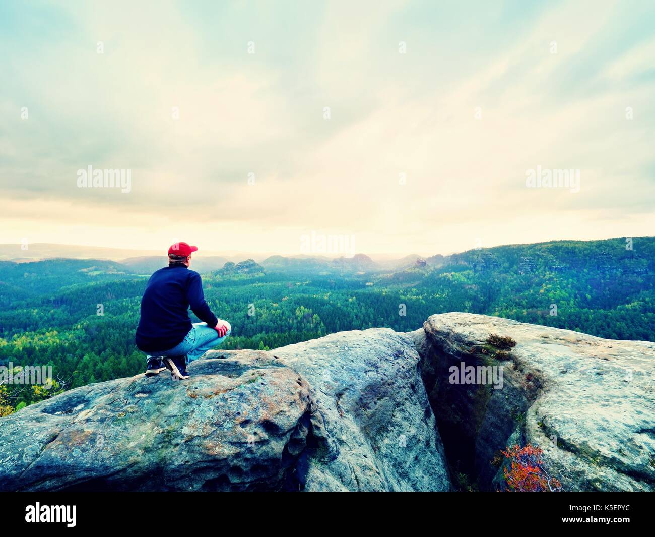Hiker man on cliff. Tourist in red cap, black sweatshirt and jeans sit ...