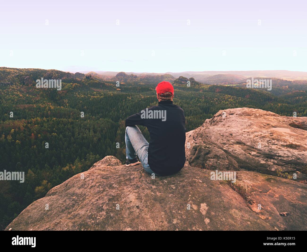 Hiker man on cliff. Tourist in red cap, black sweatshirt and jeans sit ...