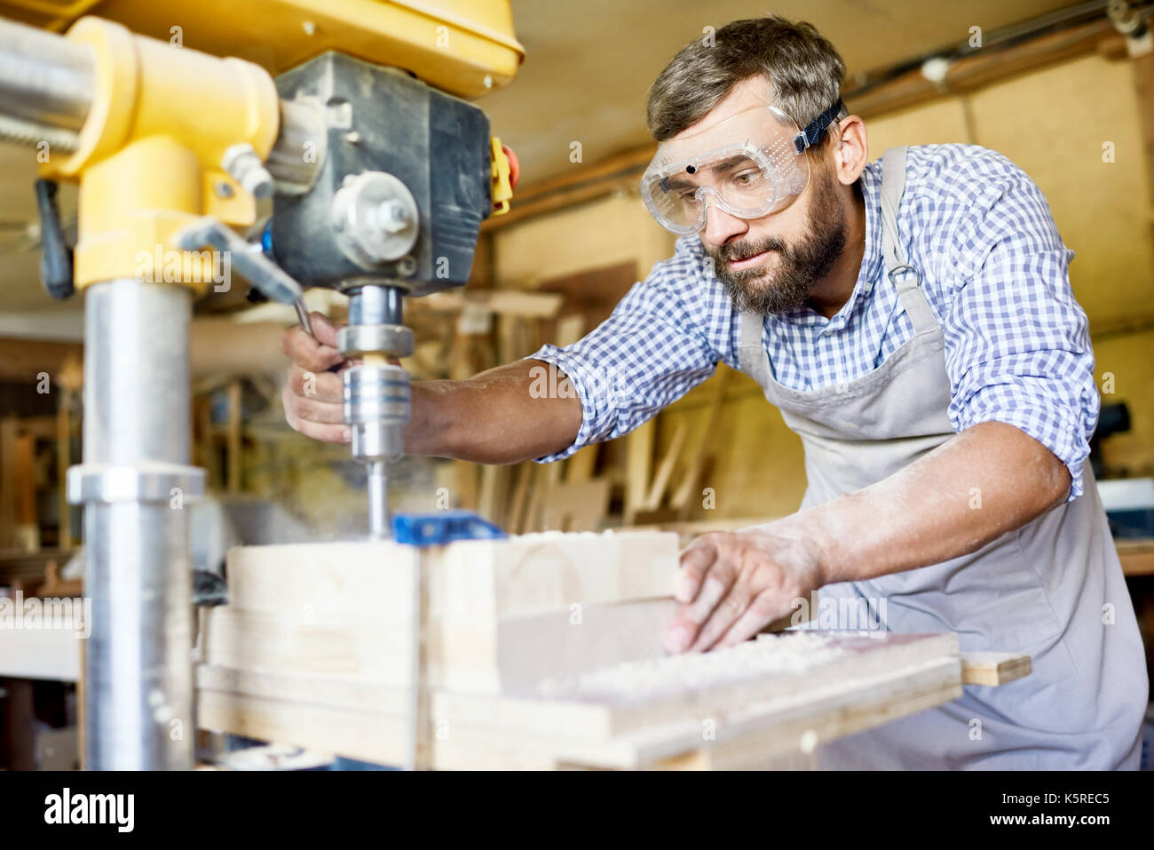 Handsome bearded carpenter wearing safety goggles and apron using drill ...
