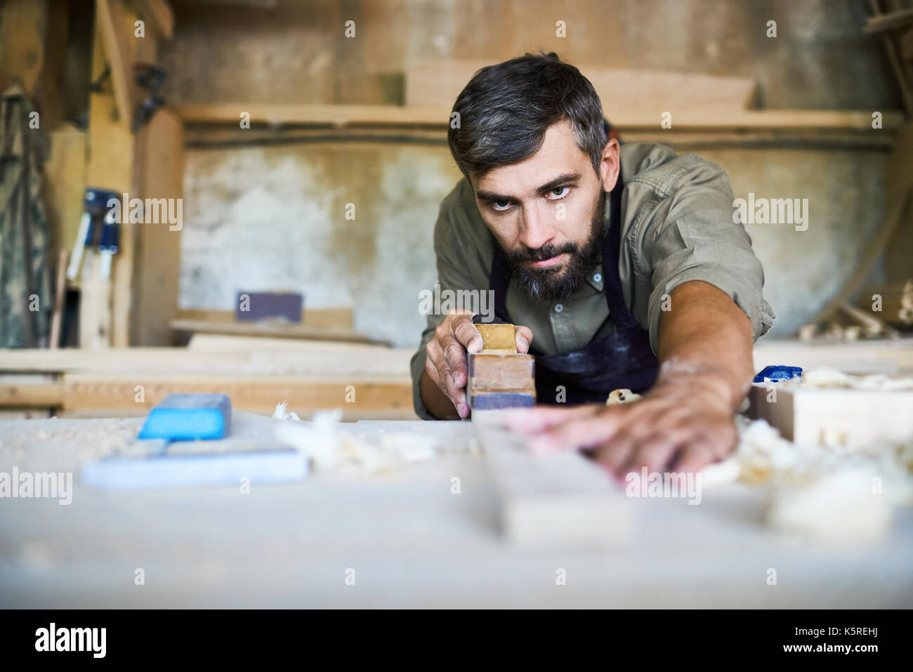 Concentrated bearded craftsman wearing apron smoothing plank with ...