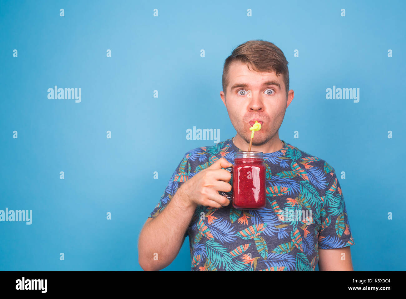 Funny young man drinking strawberry smoothie on blue background with ...