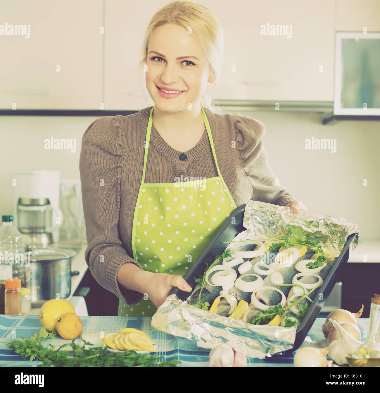 Happy smiling blonde woman cooking fish with lemon in kitchen at home ...