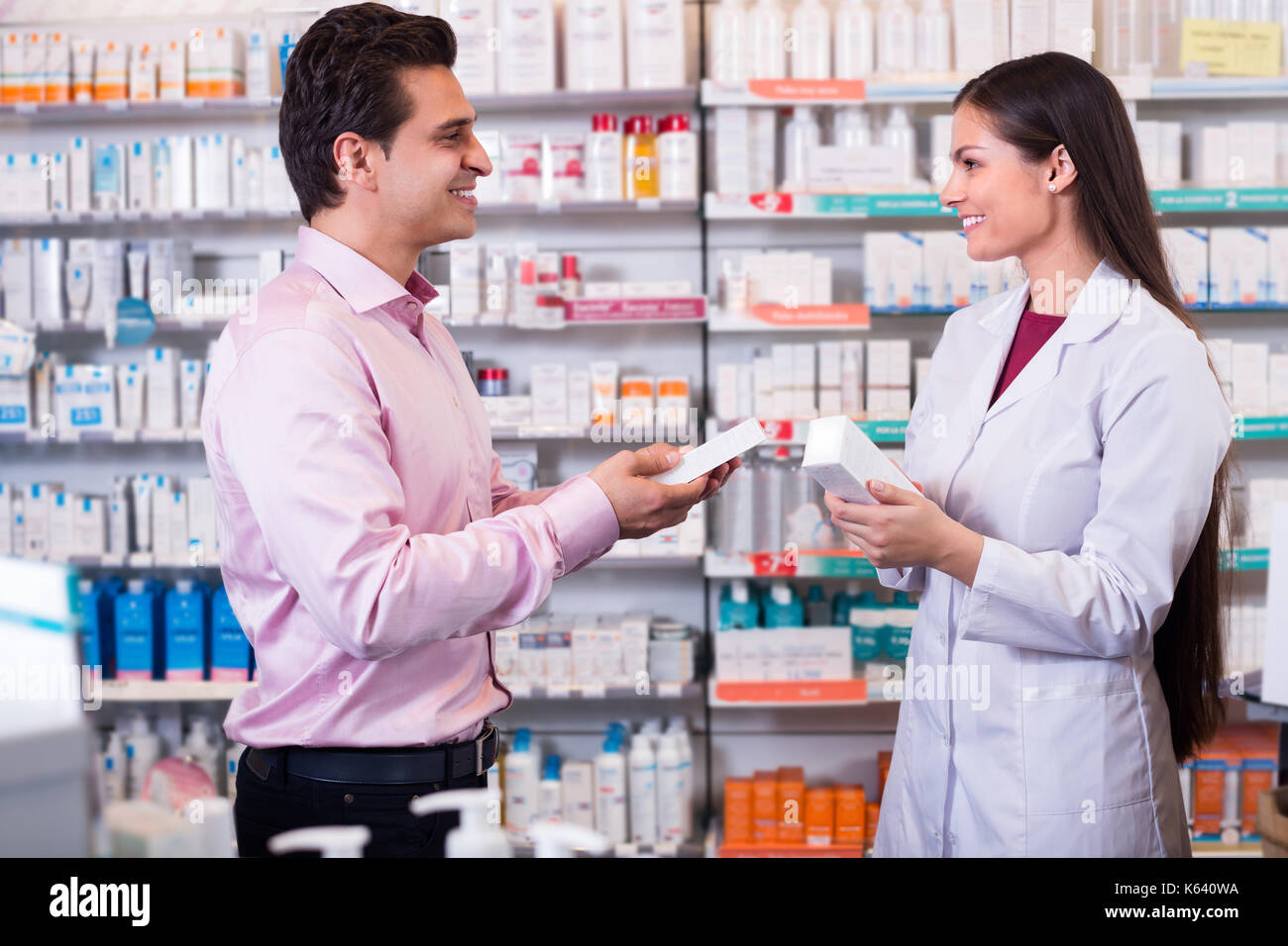 Young smiling female pharmaceutist in drugstore helping customer to ...