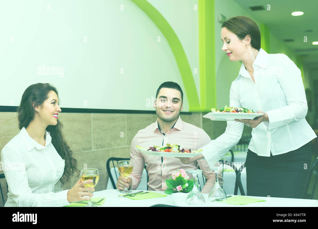 Positive smiling female waiter with plates serving visitors table in ...