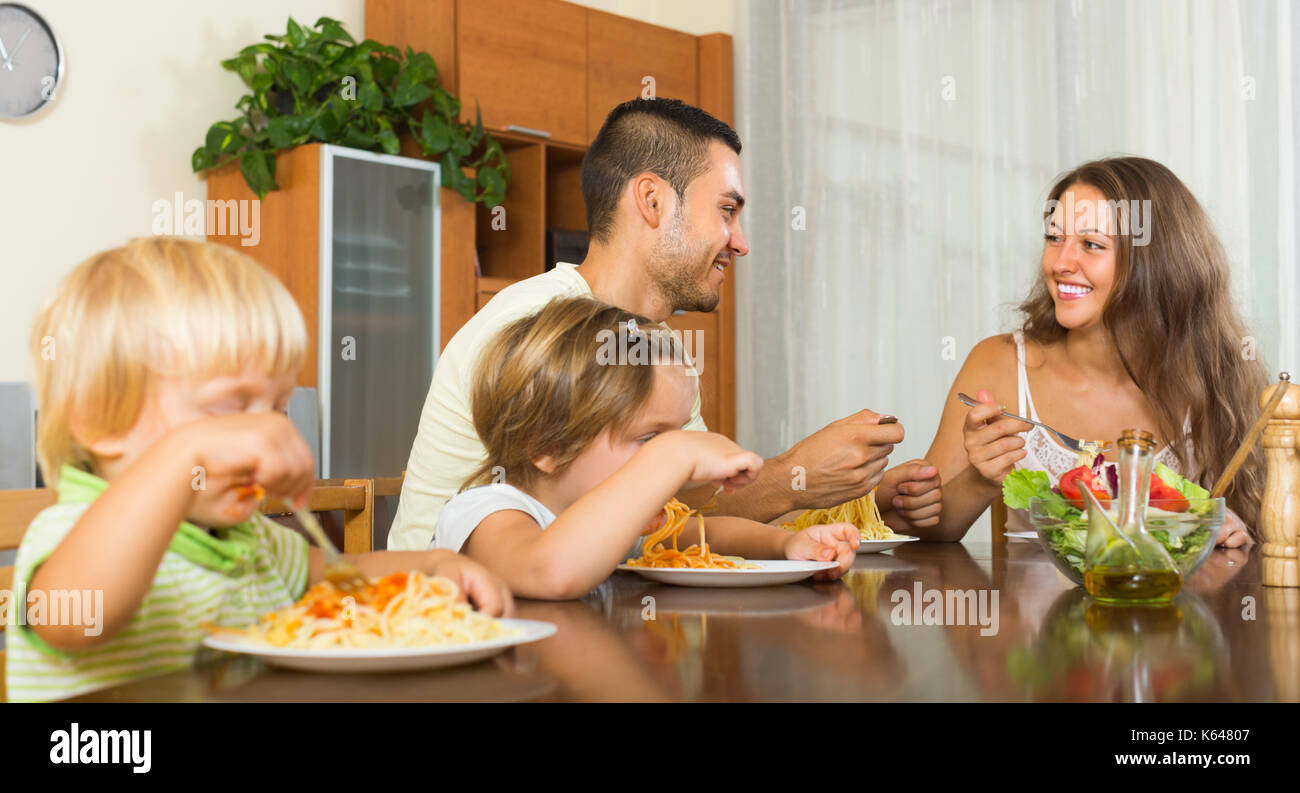 Positive young family of four eating spaghetti at home interior. Focus ...