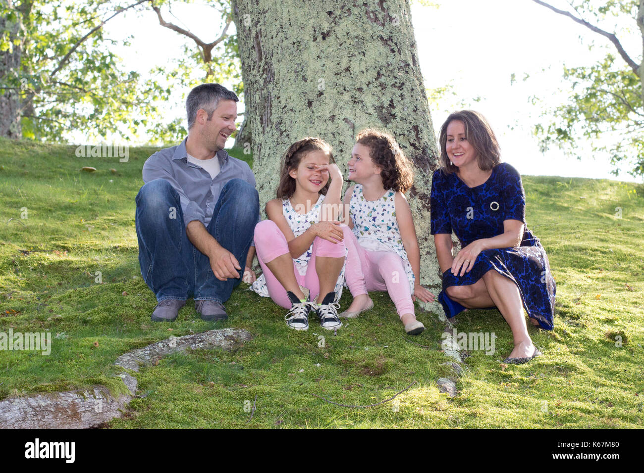 young family sits by the base of a tree outside, laughing and giggling ...