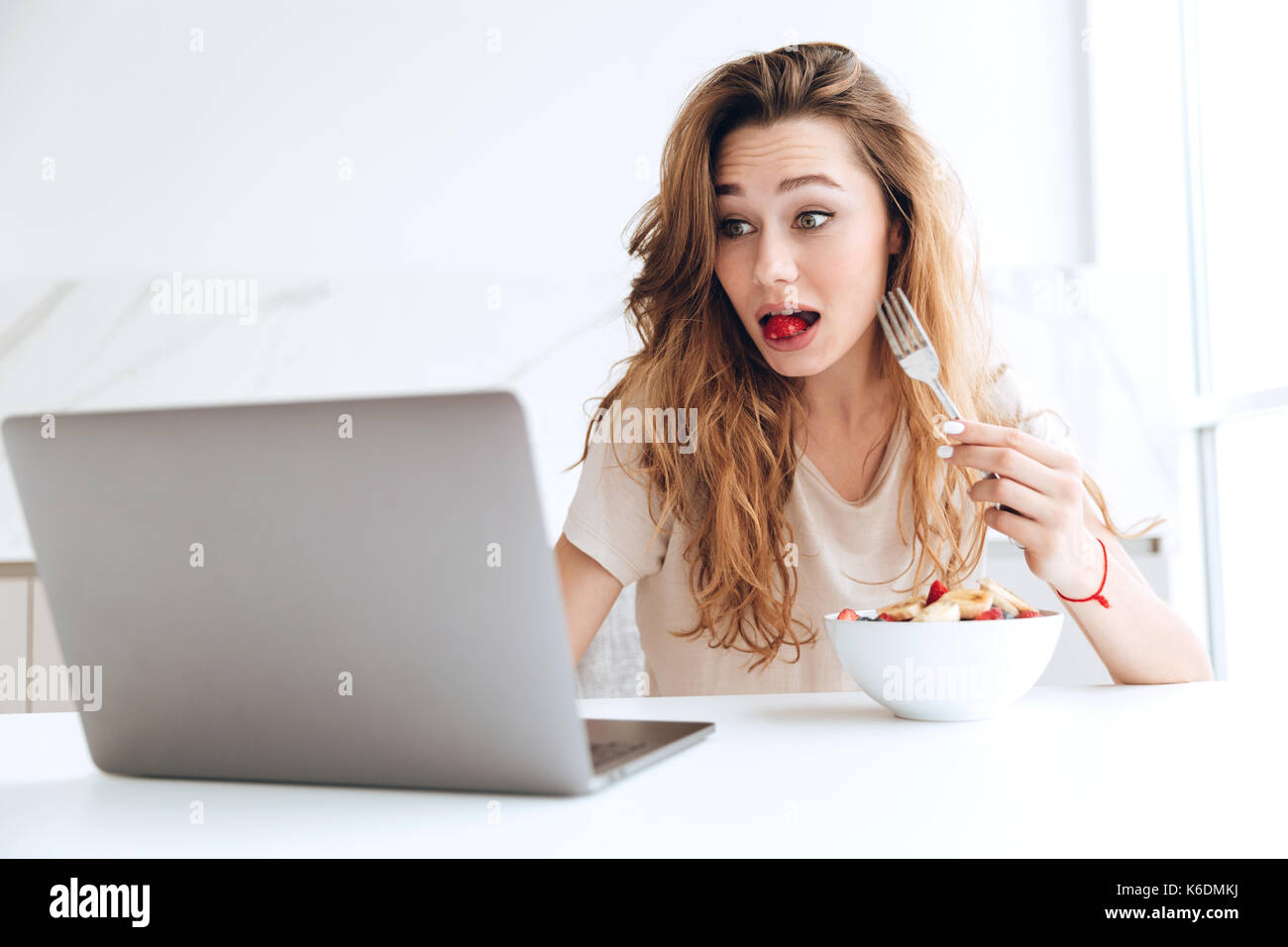 Surprised young woman eating fruits and using laptop computer at home ...