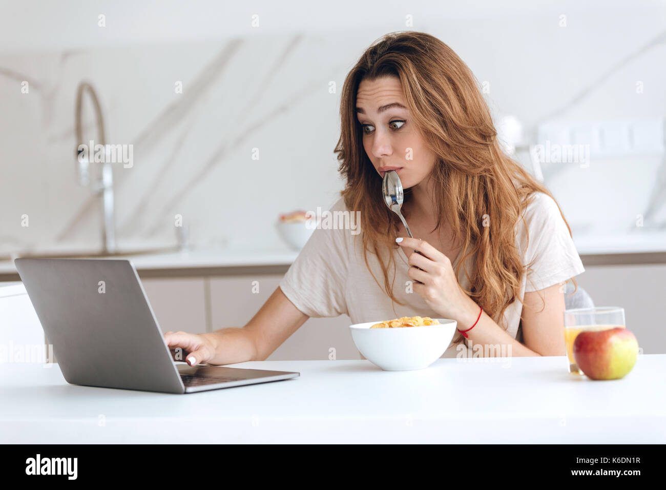 Thoughtful young woman eating breakfast and using laptop computer in ...