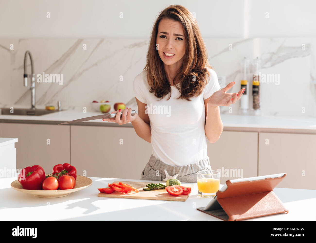Confused young woman holding knife and shrugging shoulders while ...