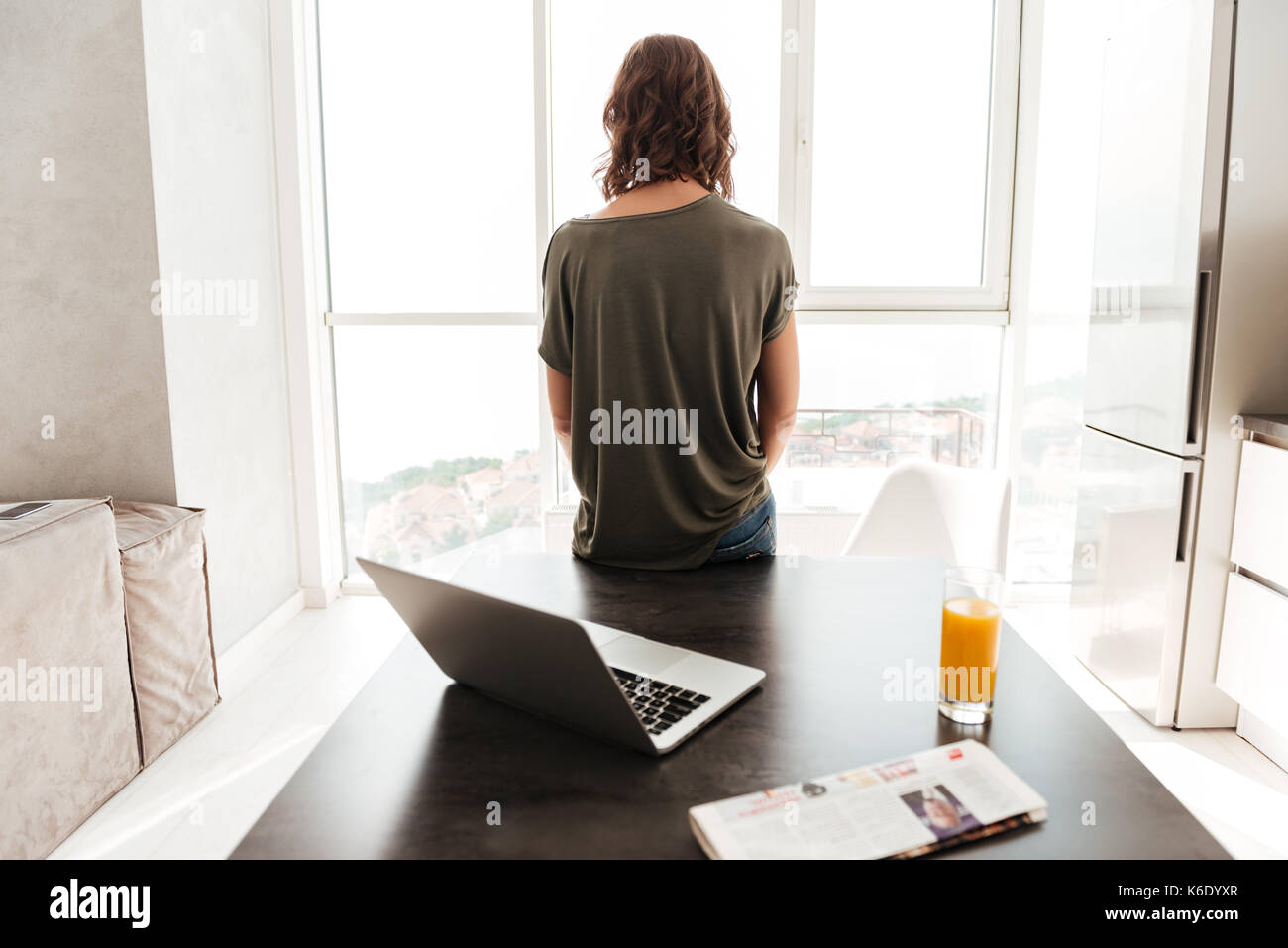 Back view of casual woman standing near table and looking at window ...