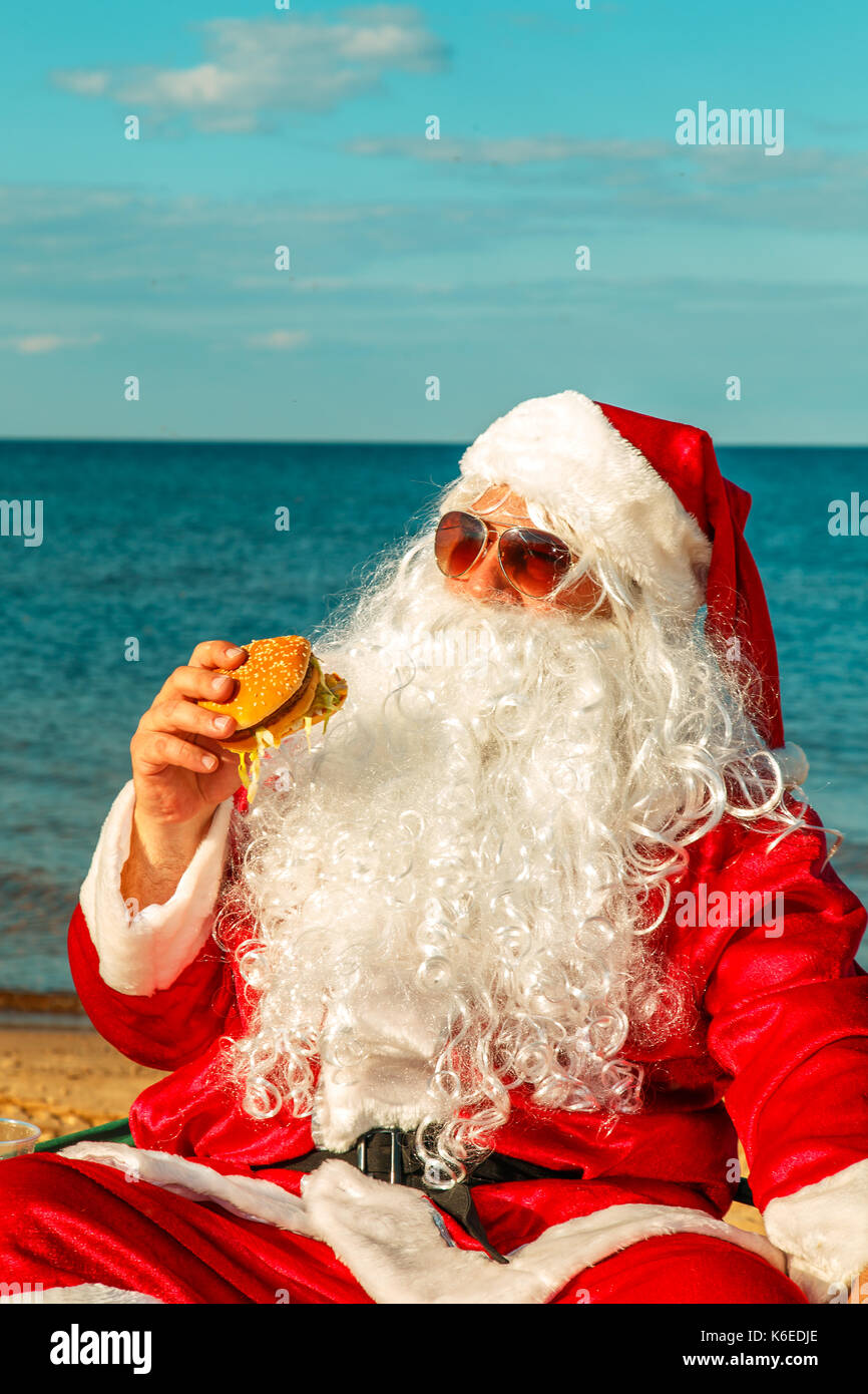 Santa Claus on the beach eating a hamburger. The concept of unhealthy ...