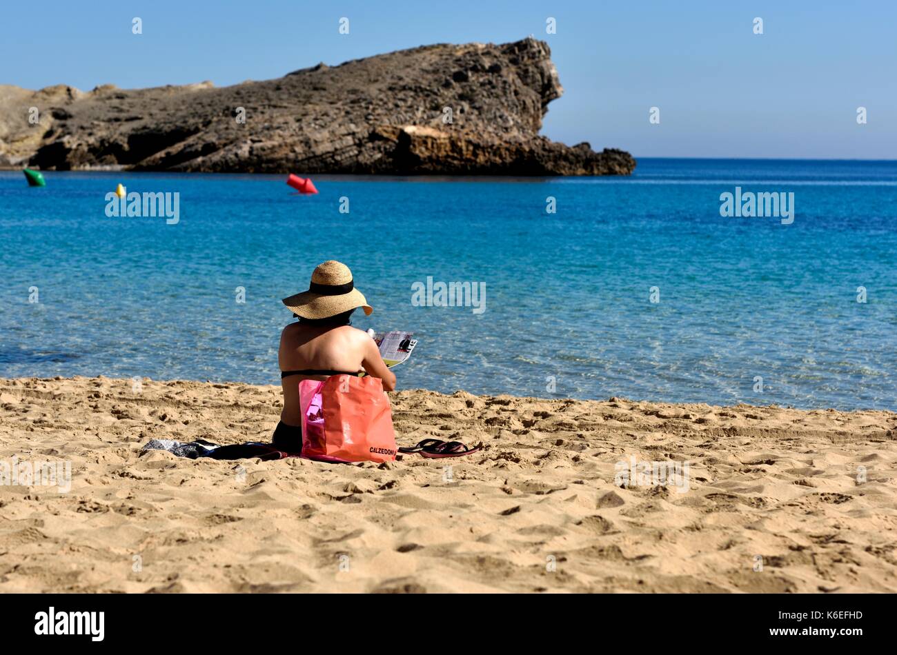 A lone woman sitting on a beach reading a magazine wearing a sun hat ...