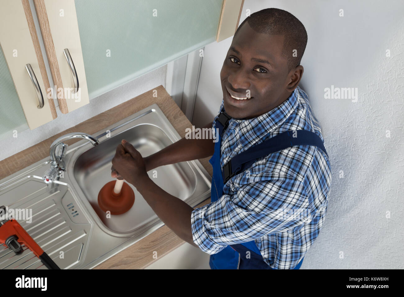 Young Happy African Plumber Using Plunger To Unclog Kitchen Sink Stock
