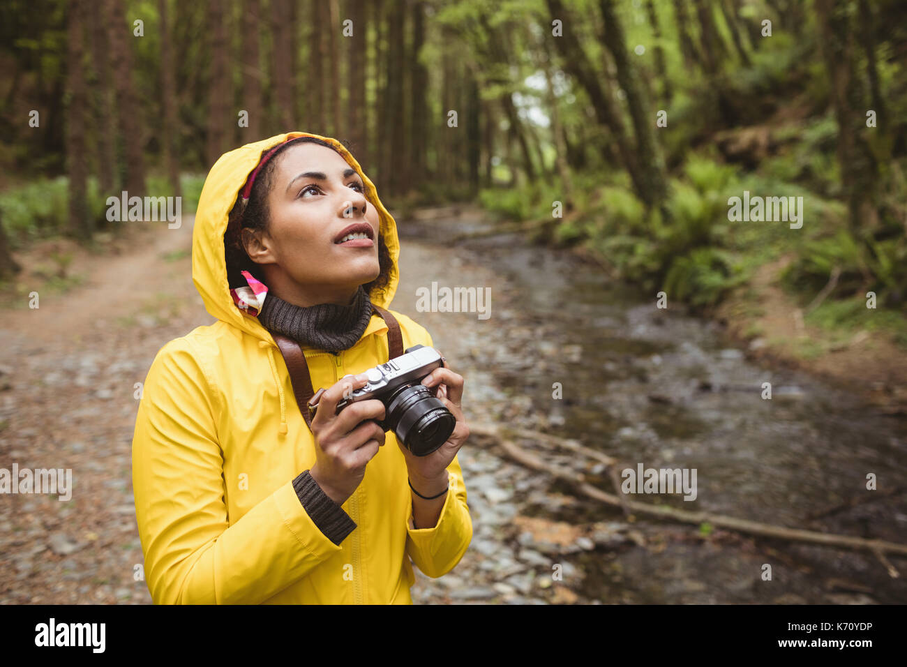 Young woman with camera looking up while standing by stream at forest ...