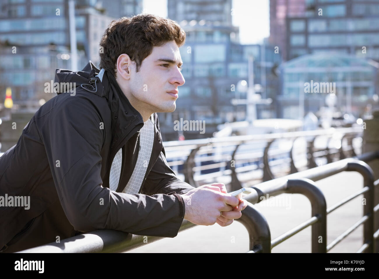 Thoughtful young man holding phone while leaning on railing against ...