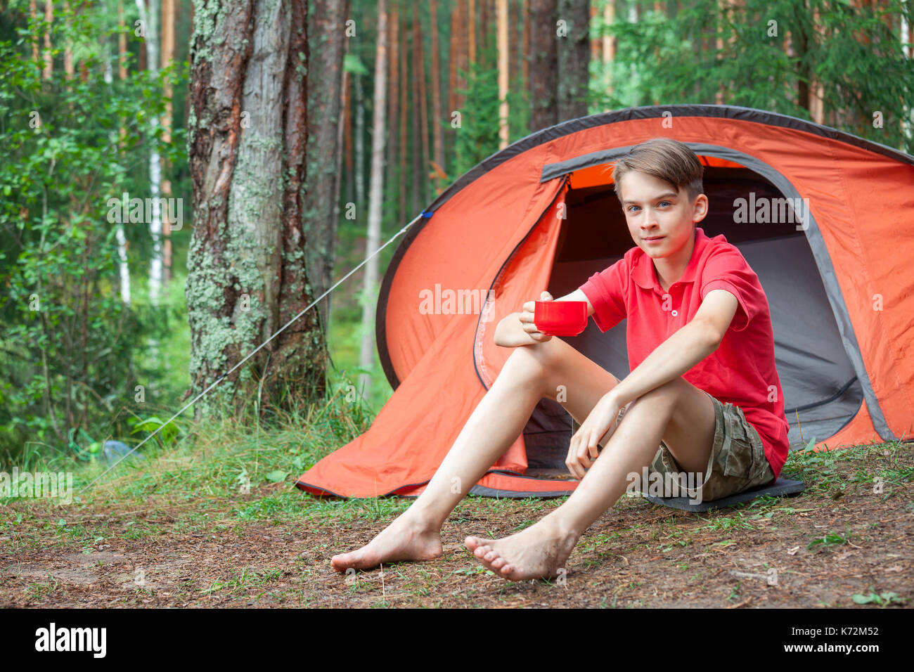 Happy teen boy sitting at a camping tent in a summer forest holding a ...