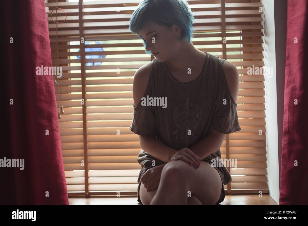 Thoughtful young woman looking down while sitting at window sill in ...
