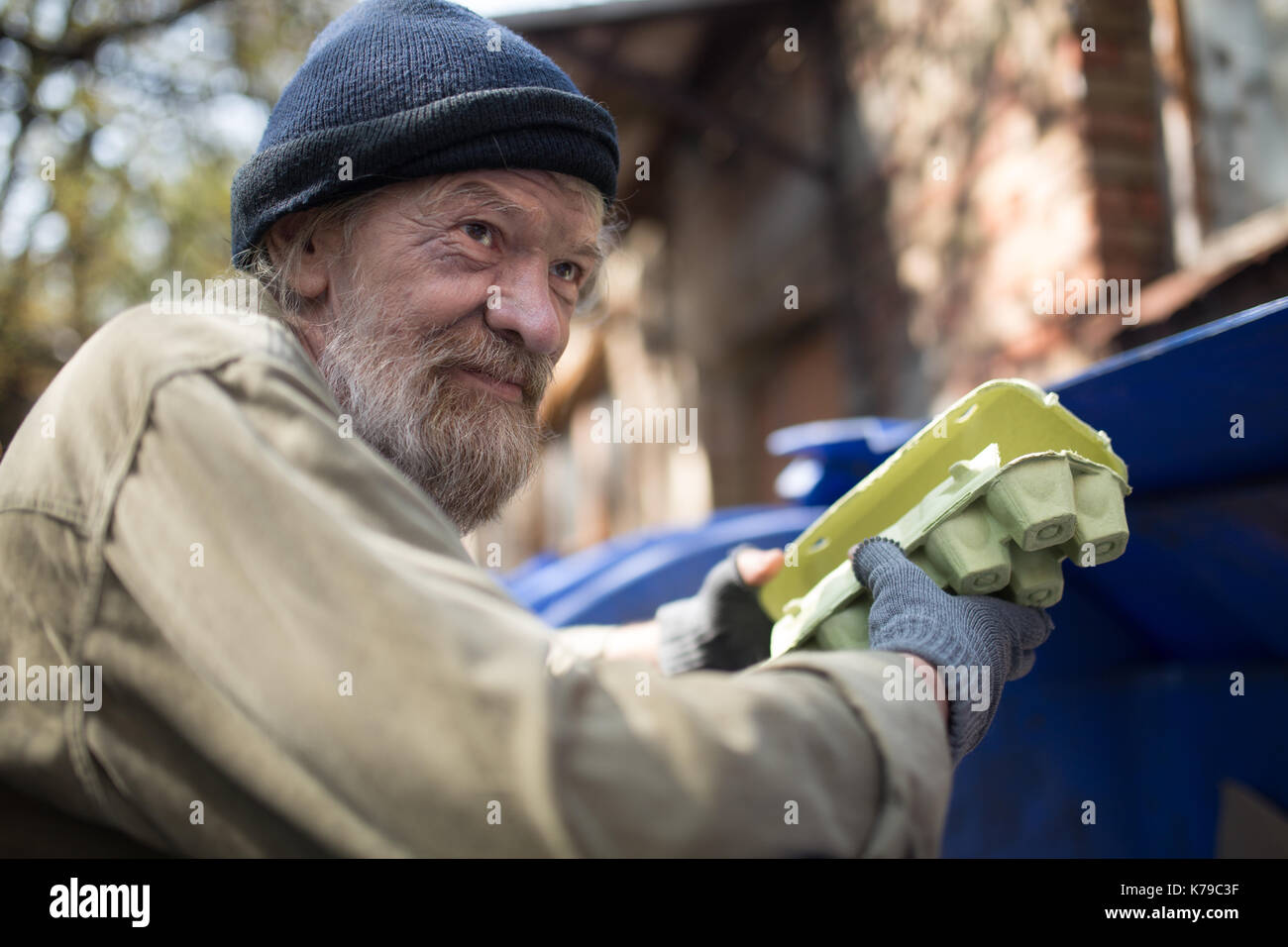 Dirty homeless man holding packing for eggs, standing by the trash can ...