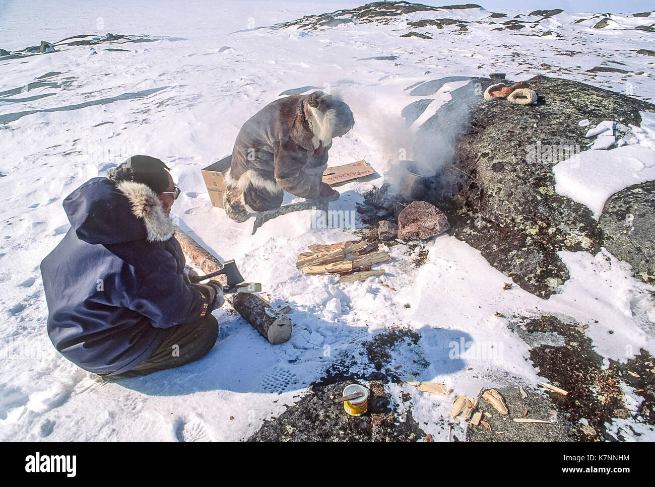 Inuit elders, 60s, build a fire to brew tea in the open tundra. One is ...