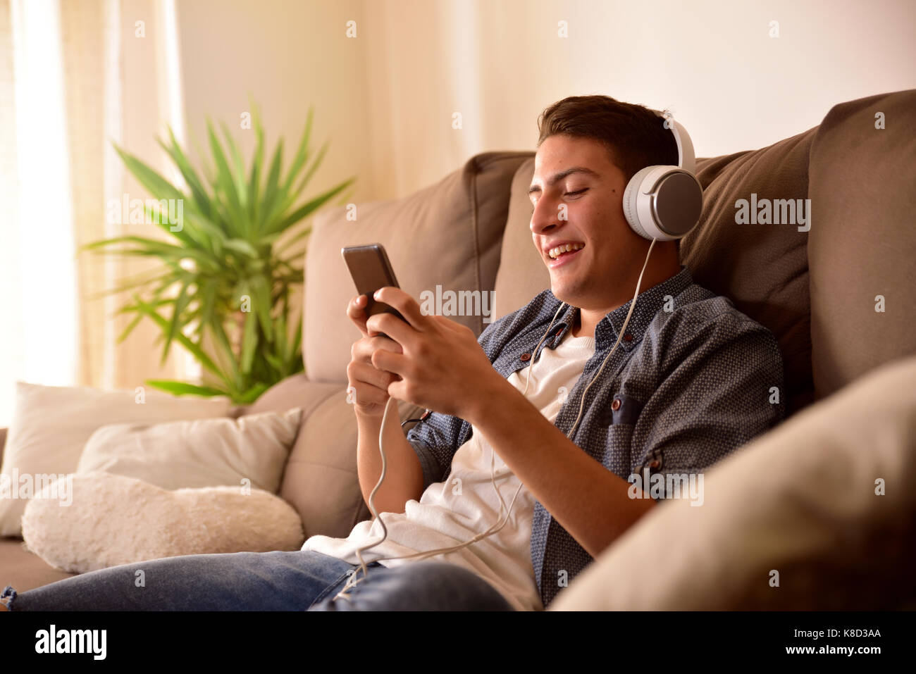 Teenager using a mobile phone with headphones sitting on a sofa at home ...