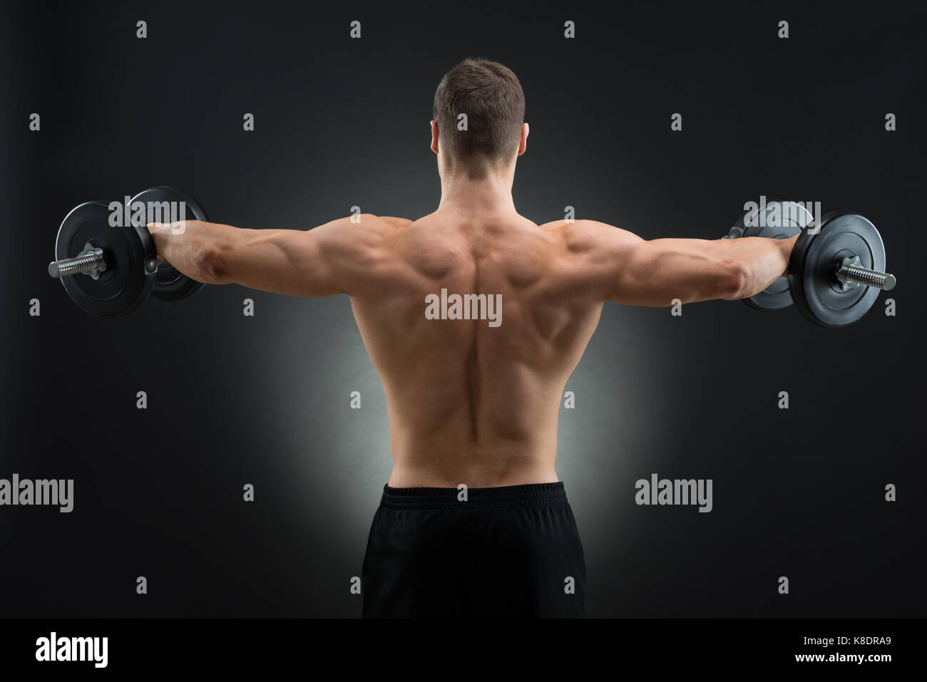 Rear view of strong man lifting dumbbells against black background ...
