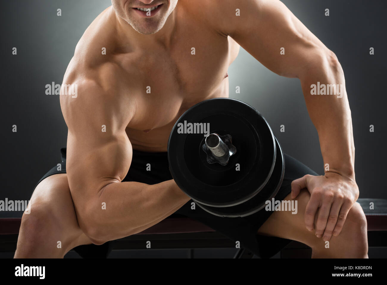 Midsection of muscular man lifting dumbbell while sitting on chair ...