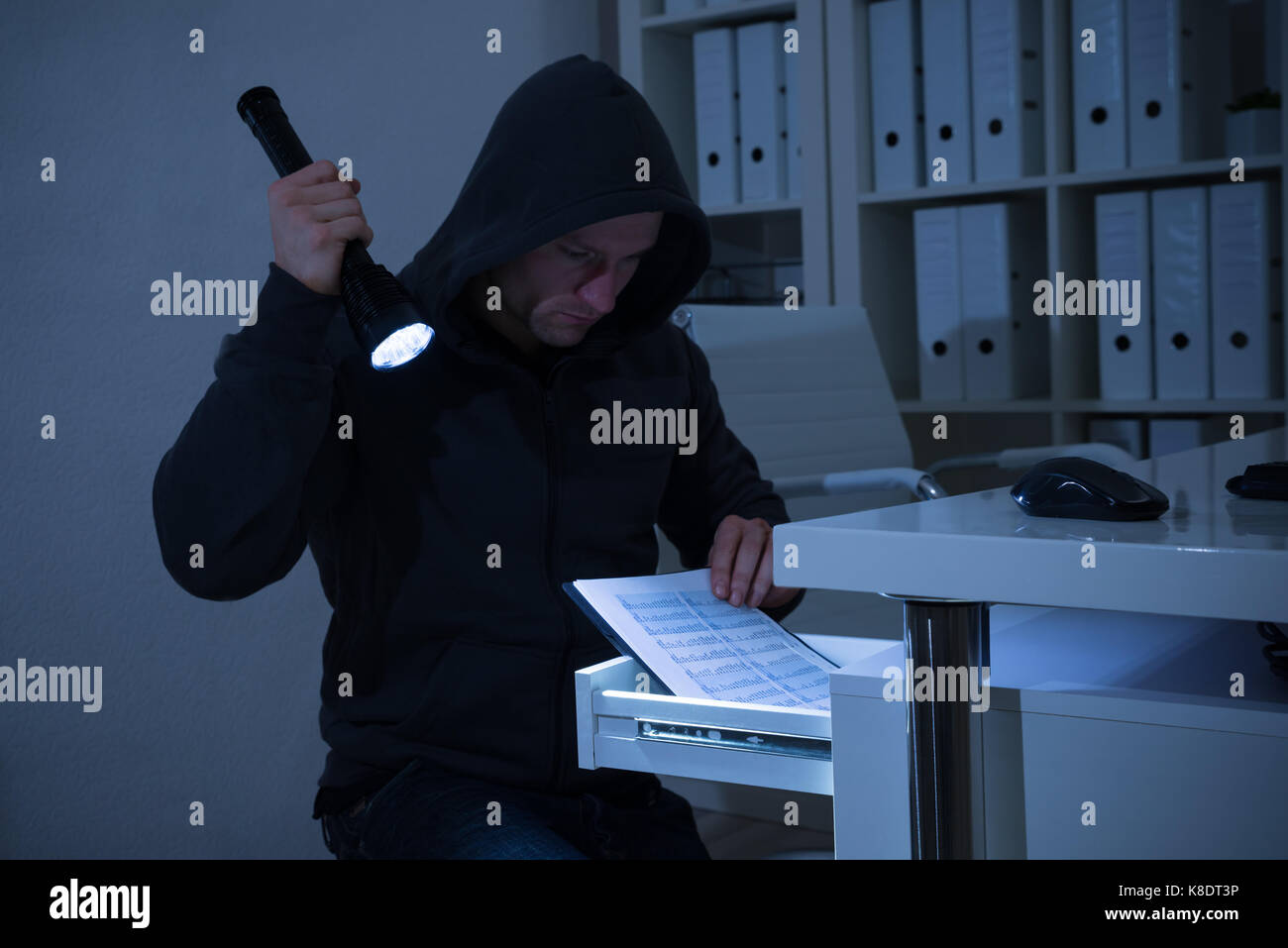 Robber with flashlight searching for documents in drawer at office ...