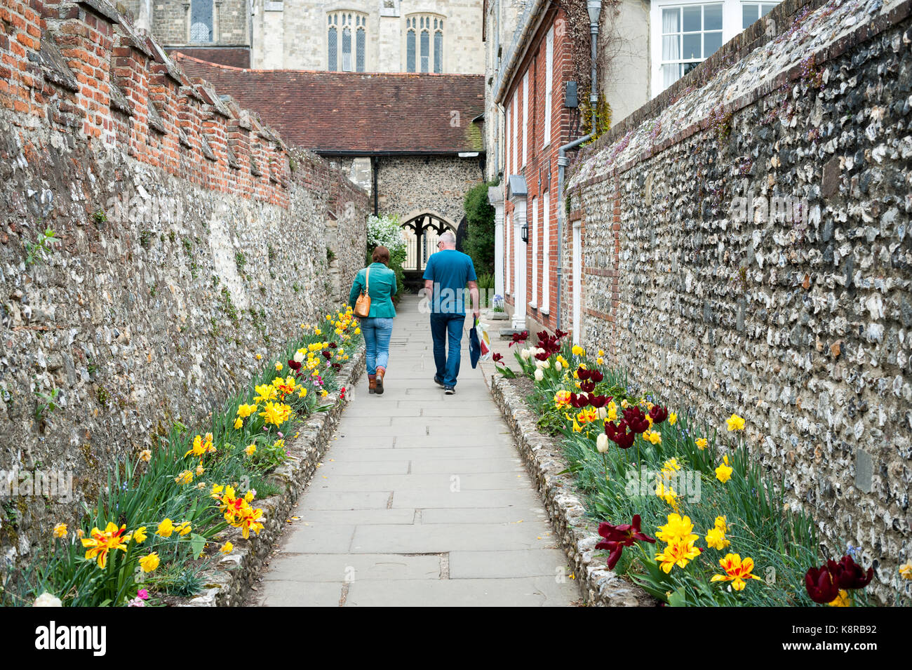 Two people pass old flint walls and colourful flowers as they walk ...
