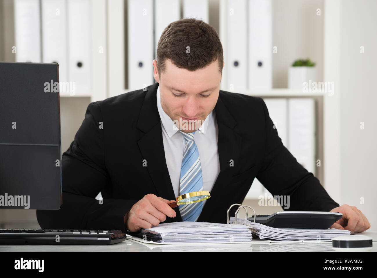 Male accountant analyzing invoice with magnifying glass at desk in ...