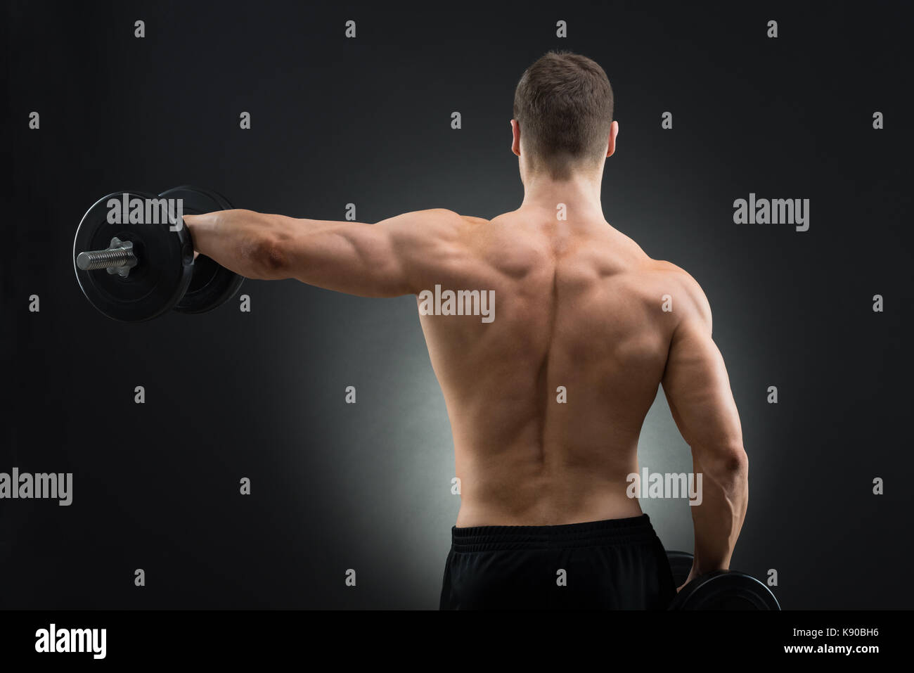 Rear view of muscular man lifting dumbbell while standing against black ...