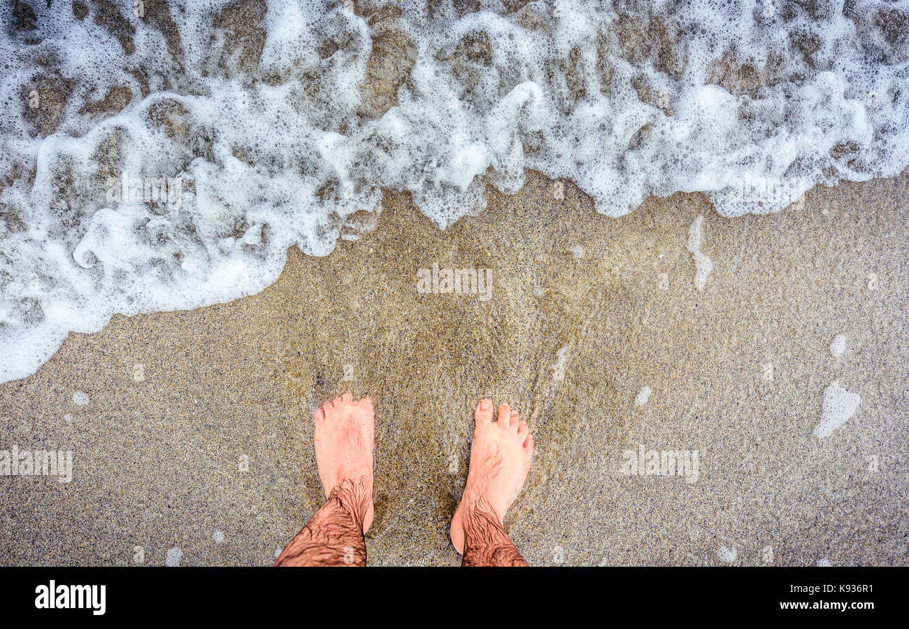 Person standing in ocean sea waves on sandy beach. Barefoot legs ...