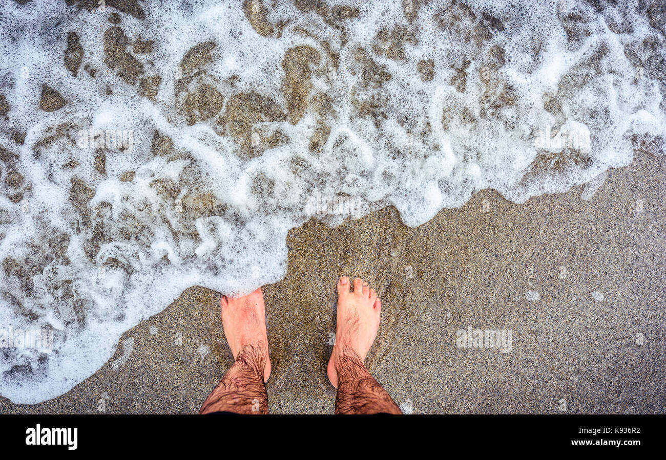 Person standing in ocean sea waves on sandy beach. Barefoot legs ...