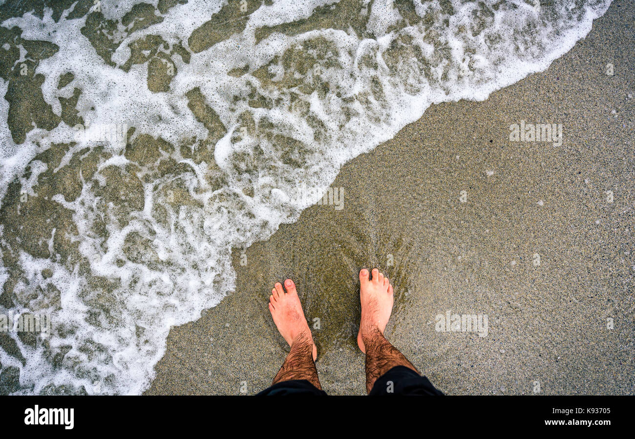 Person standing in ocean sea waves on sandy beach. Barefoot legs ...