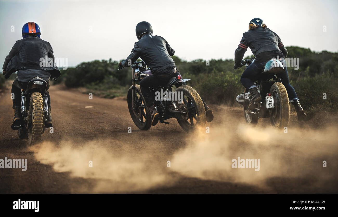 Rear view of three men riding cafe racer motorcycles along dusty dirt ...