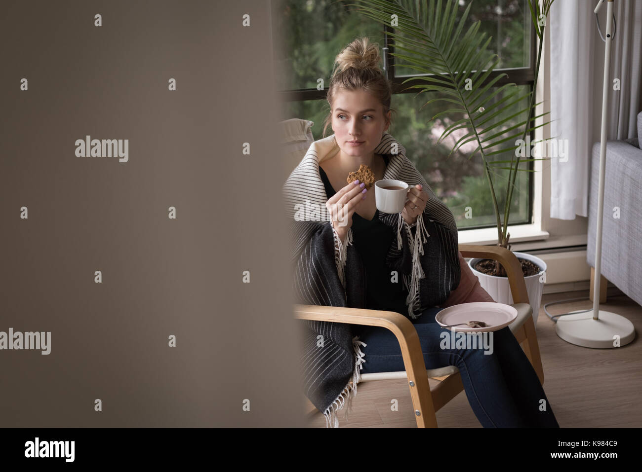 Thoughtful woman having breakfast while sitting on chair by window at ...