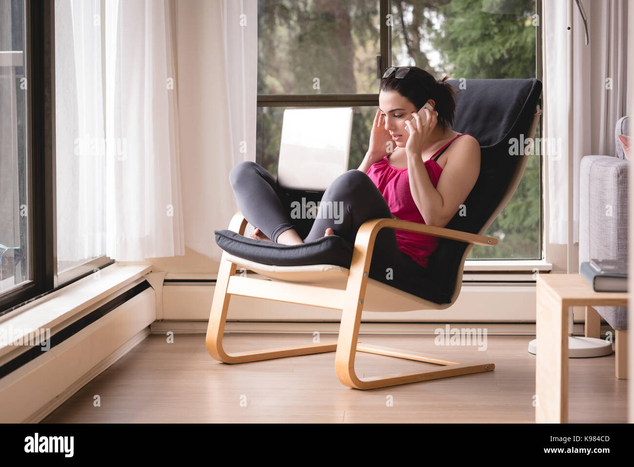Young woman calling while sitting on chair by window at home Stock ...