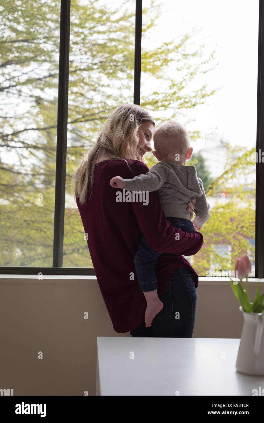 Rear view of woman carrying son while standing by window at home Stock ...