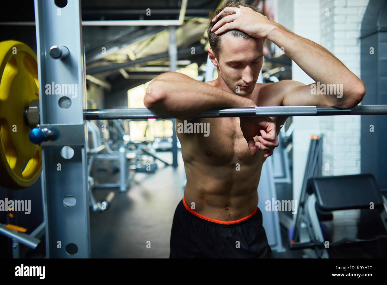 Exhausted young athlete with bare muscular torso leaning on barbell ...