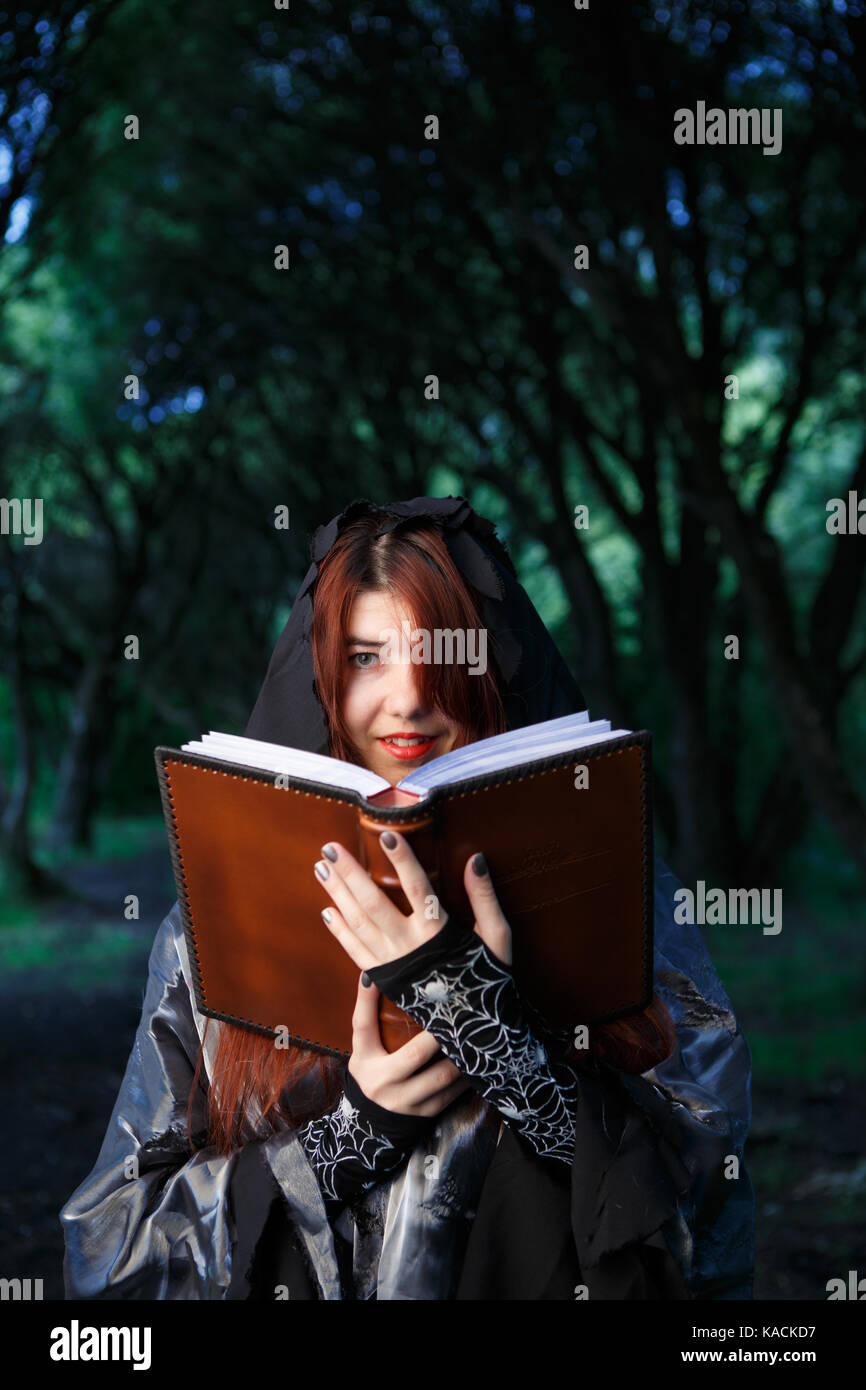 Picture of witch in black hood with book in hands at dark evening ...