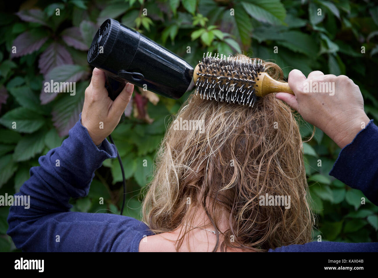 Woman drying her long hair with a hair dryer, a foliage in the blurred ...