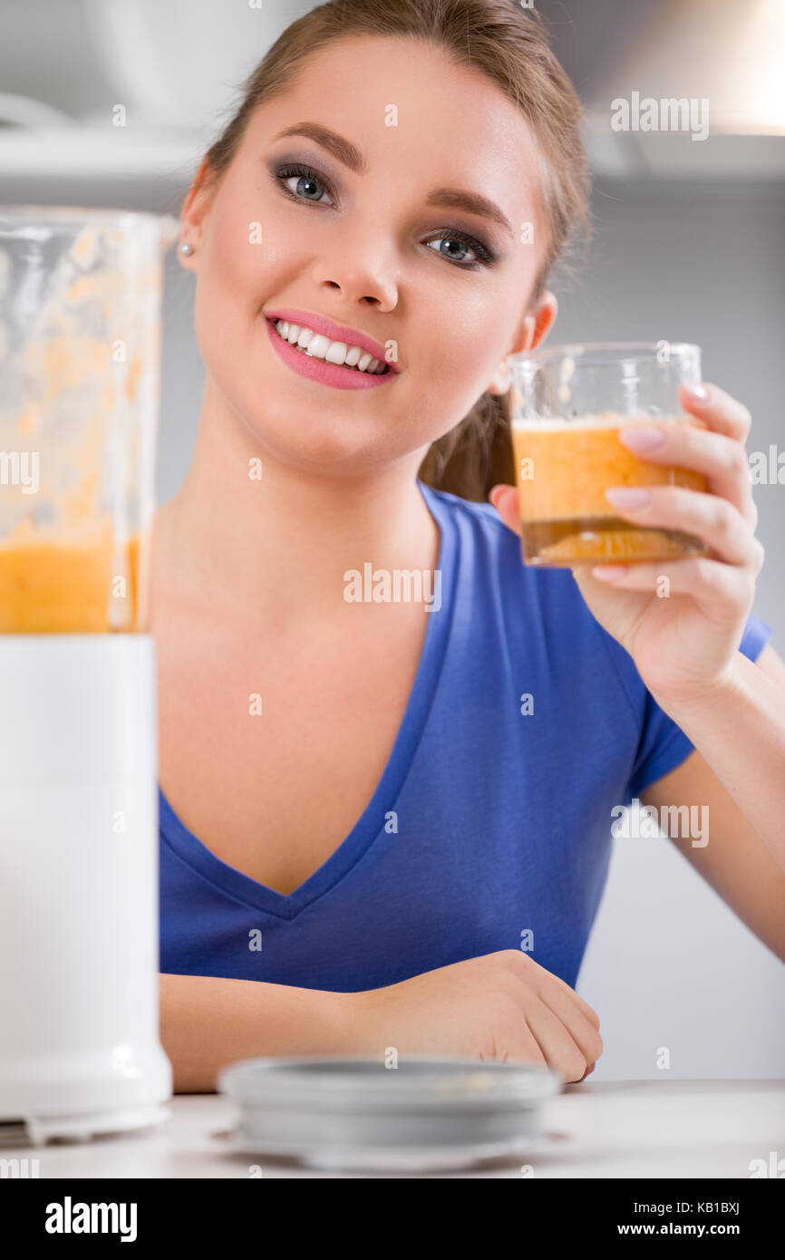 Beautiful woman using a blender while holding a drink in the kitchen ...