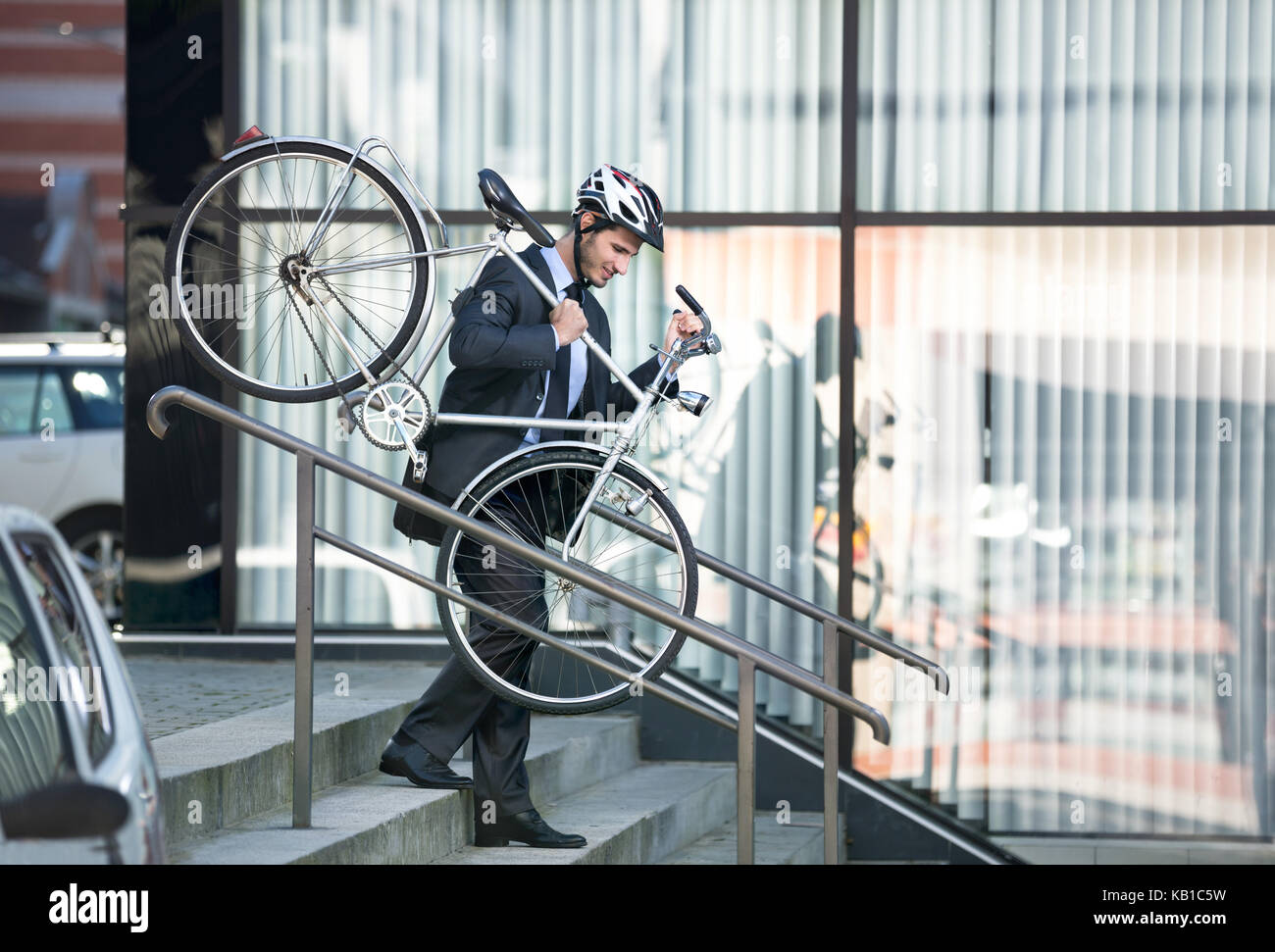 businessman with a cycle helmet on head carrying his bike down steps ...