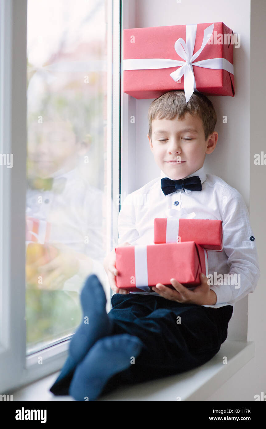 happy little boy in bow tie sitting by window with a gift box on his ...