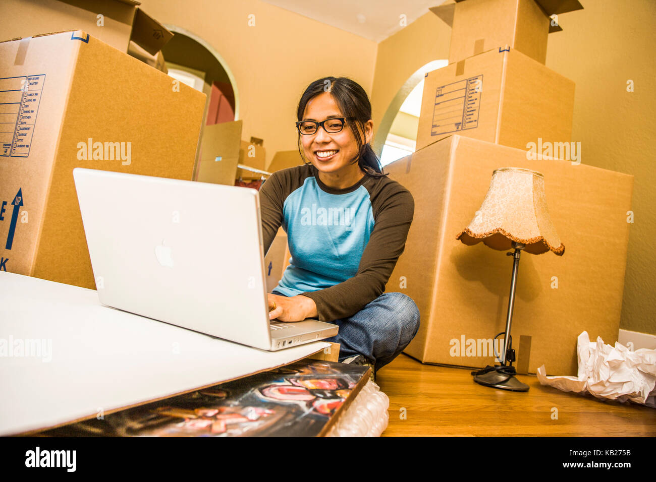 A smiling young woman sitting on the floor surrounded by moving boxes ...