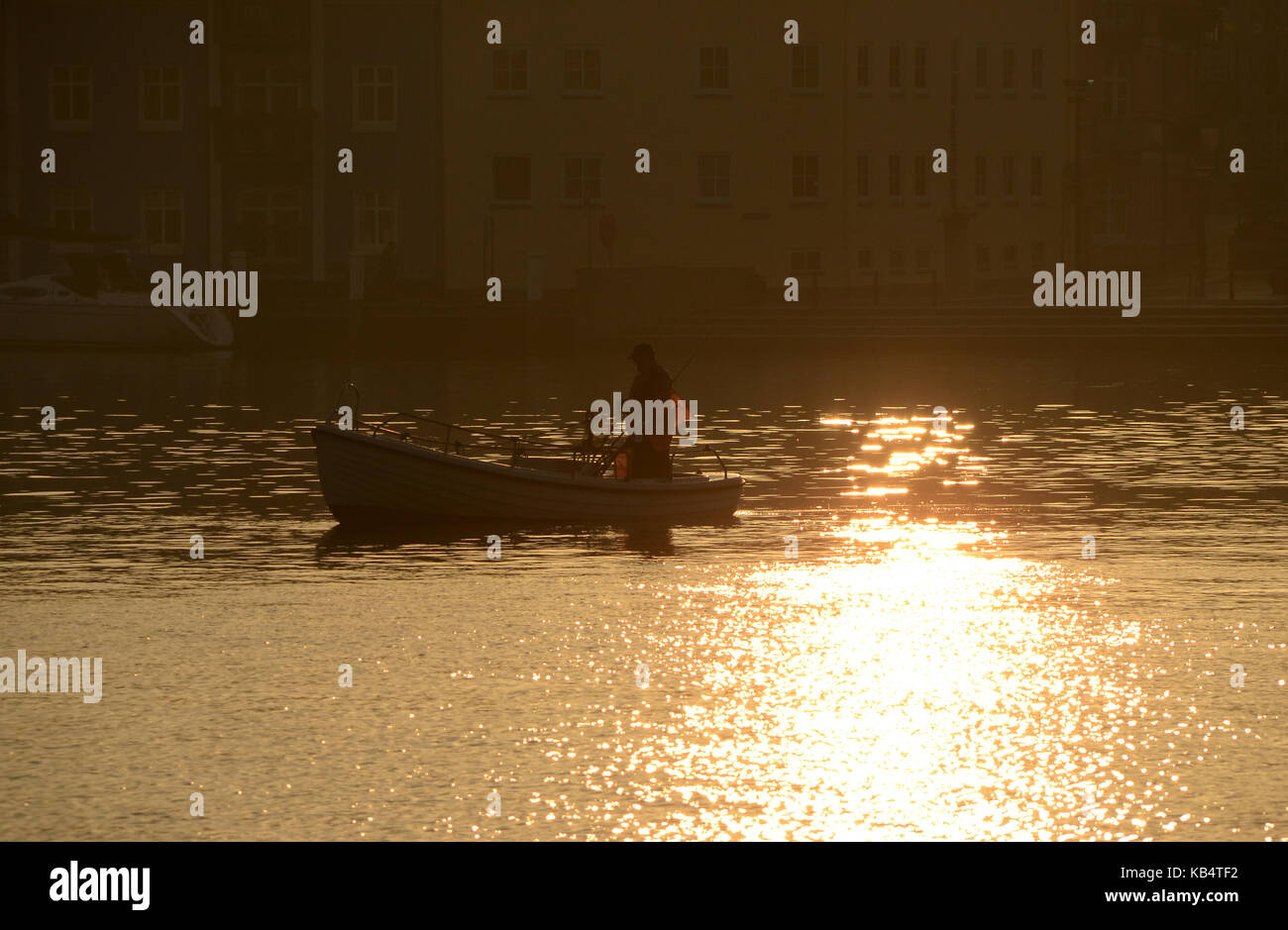Fisherman in a boat seen in harsh back light with an urban backdrop ...